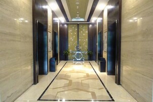 Um hall de elevadores moderno com piso de padrão geométrico, uma luminária central e uma parede refletora.