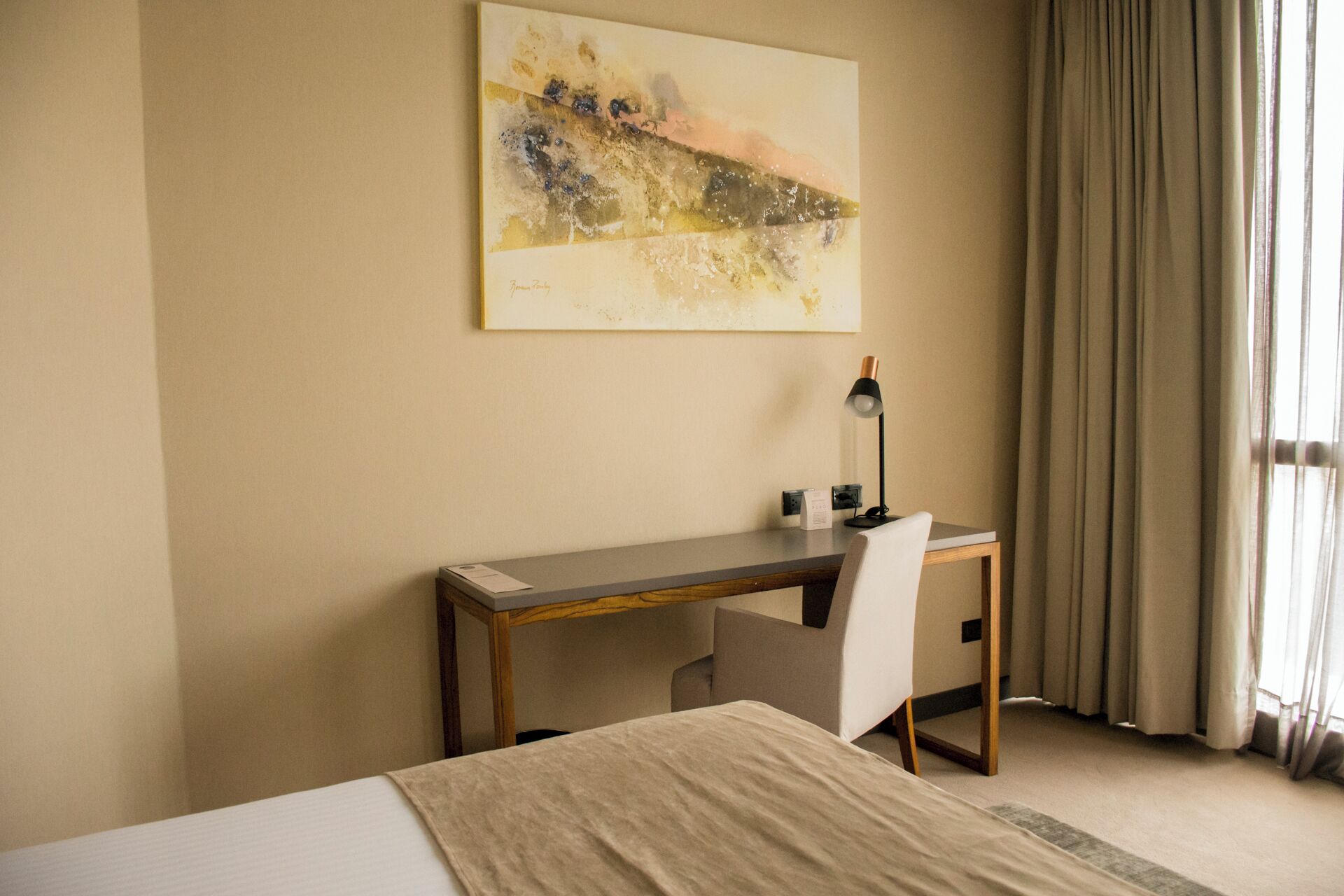 Quarto de hotel com cama, escrivaninha, cadeira, abajur e um quadro na parede.
