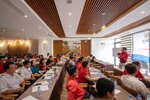 Sala de conferência com painéis de madeira, uma tela grande e participantes sentados em mesas com garrafas de água.