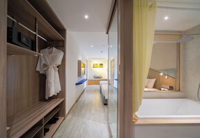Quarto de hotel moderno com closet espaçoso, cama grande, banheiro separado com chuveiro e mesa de cabeceira com abajur.