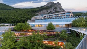 Um hotel moderno com fachada de vidro, localizado num vale verdejante com montanhas ao fundo.