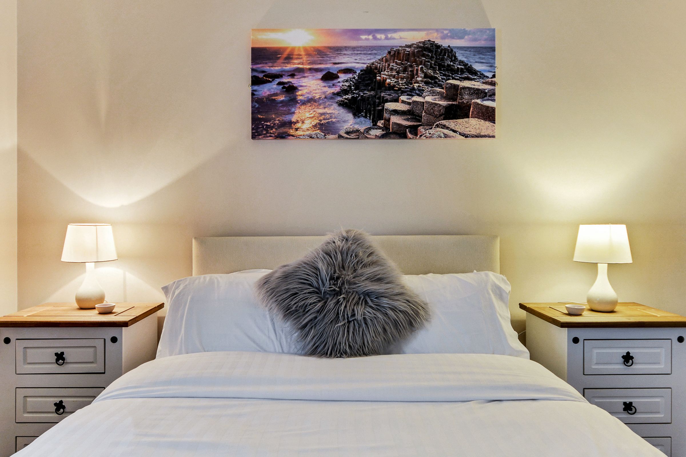 Quarto com cama, duas mesas de cabeceira com luminárias, um quadro pendurado na parede retratando uma paisagem costeira e um travesseiro macio na cama.