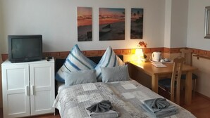 Quarto de hotel com uma cama, televisão, secretária com abajur e um espaço de refeições.