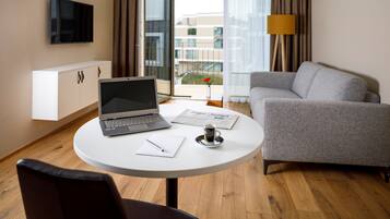 Quarto de hotel com um laptop sobre uma mesa redonda, um sofá, uma TV fixada na parede e uma janela com cortinas.