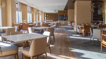 Uma sala de jantar espaçosa com várias mesas e cadeiras, grandes janelas que proporcionam muita luz natural, e uma cozinha moderna ao fundo.