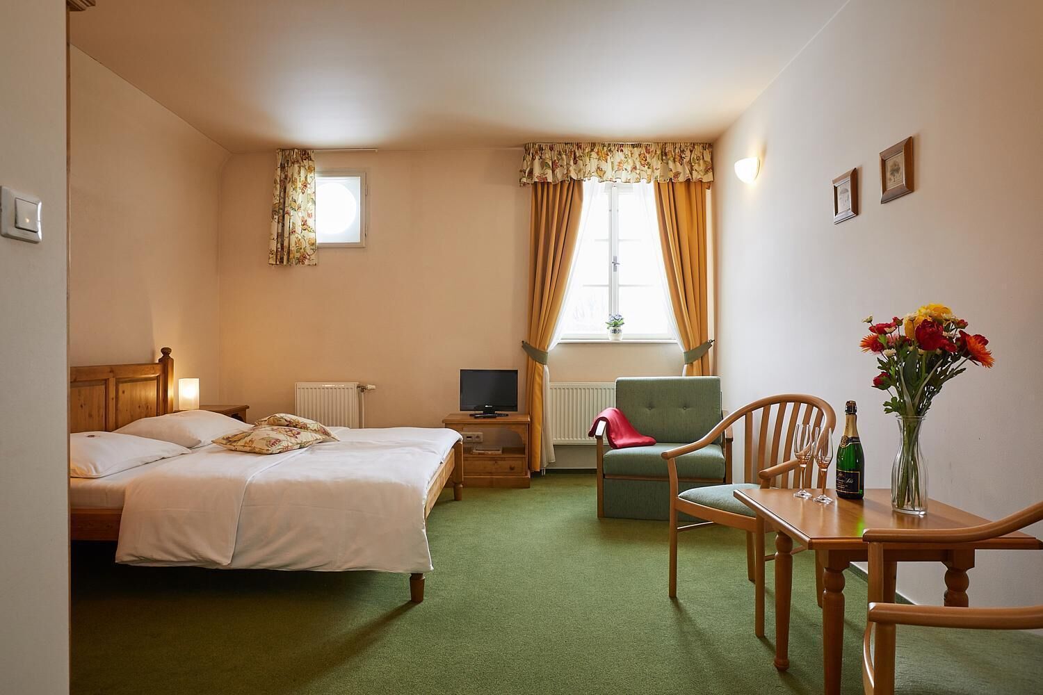 Quarto de hotel com cama, televisão, cadeira, mesa com garrafa e flores, e janela com cortinas.