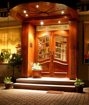 Entrada de hotel com fachada de madeira, portas duplas e um letreiro sobre as portas. Há plantas em vasos de ambos os lados da entrada.