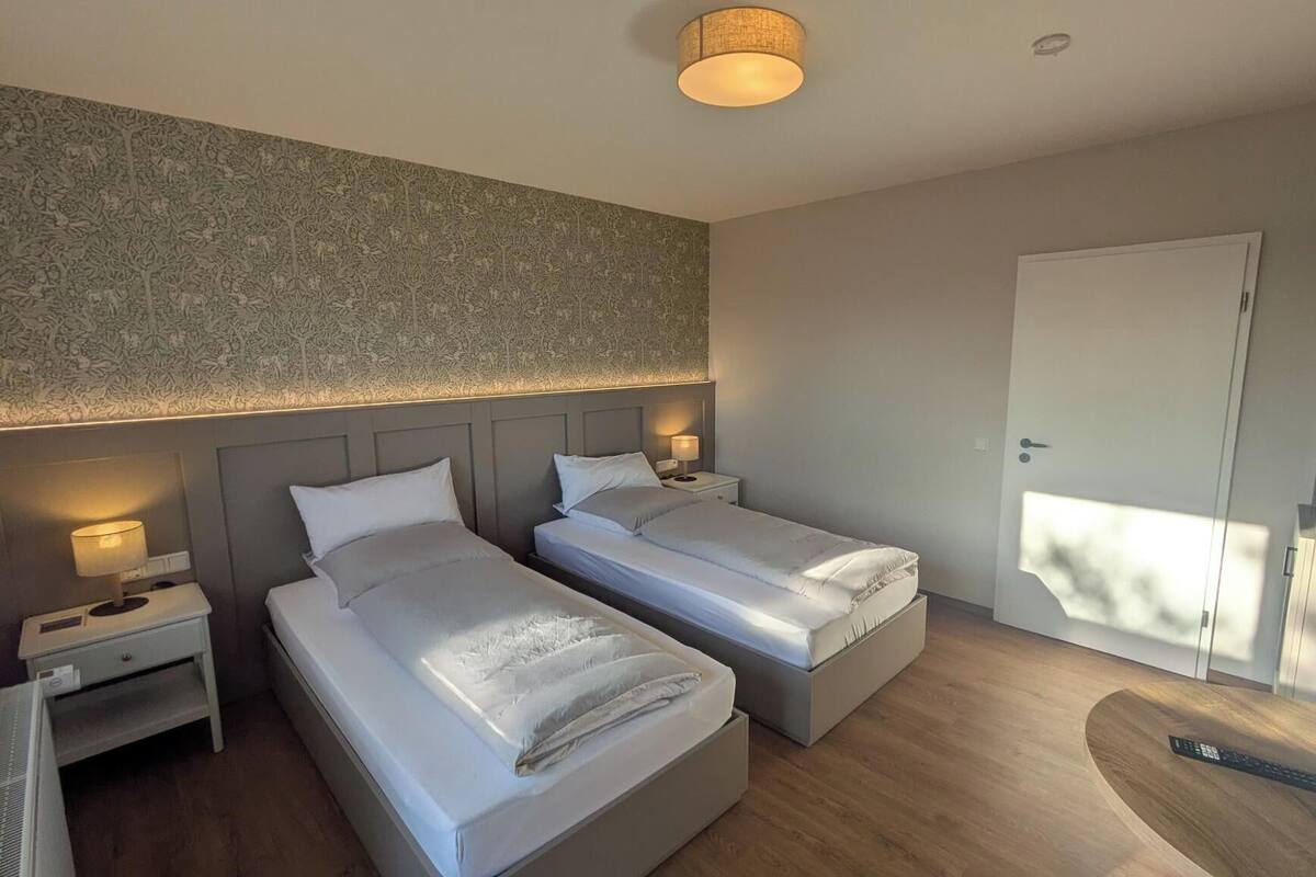 Quarto de hotel com duas camas, uma mesa de cabeceira com abajur, uma escrivaninha de madeira e uma porta.