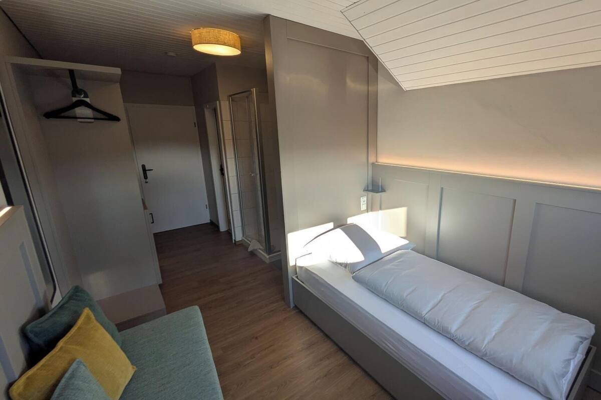 Quarto pequeno e bem arrumado com uma cama de solteiro, luminária pendente e porta que dá para outro cômodo.