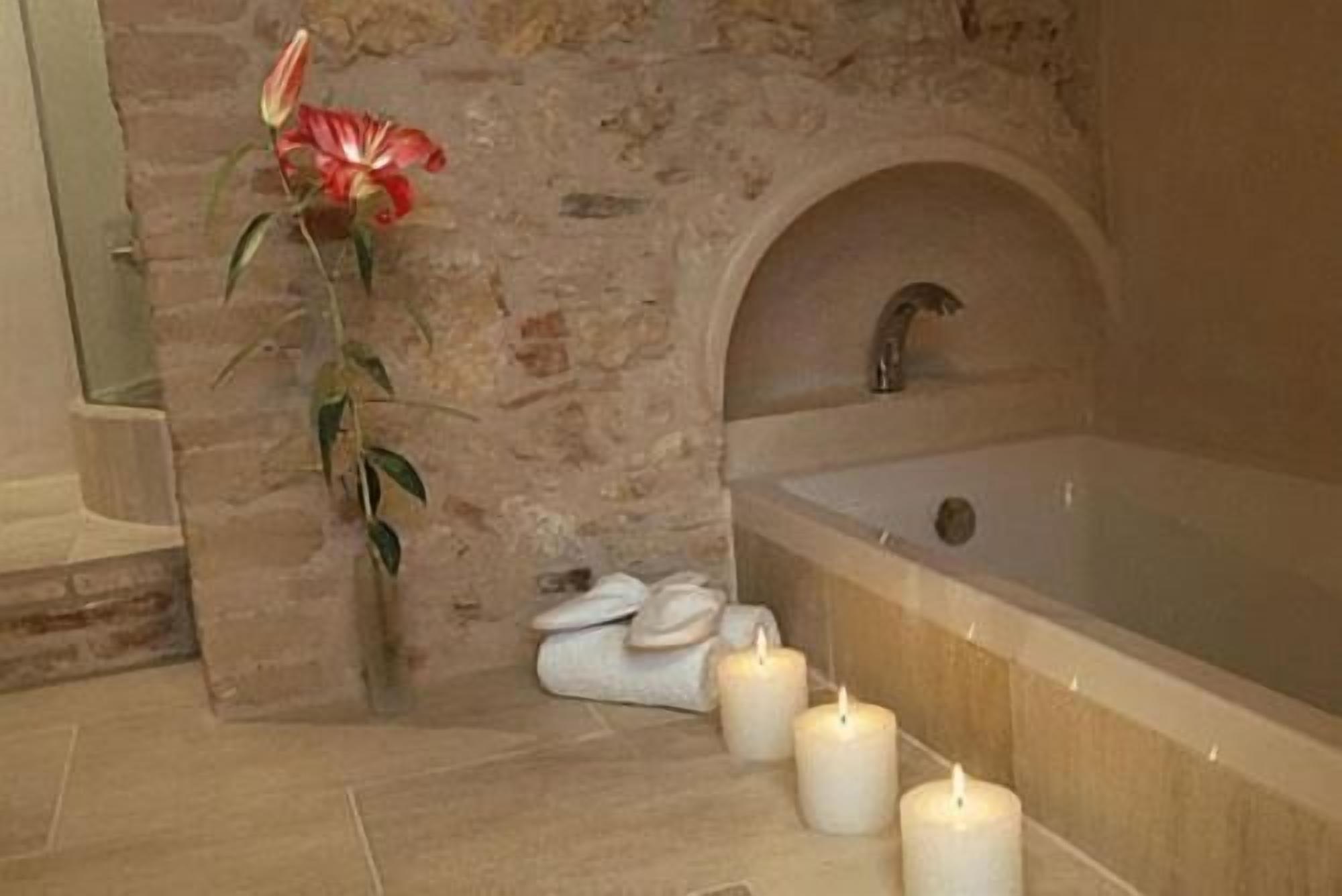 Casa de banho com paredes de pedra, banheira, velas e uma flor vermelha.