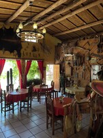 Uma área de refeições rústica com mesas e cadeiras de madeira, um candelabro e diversos objetos pendurados.