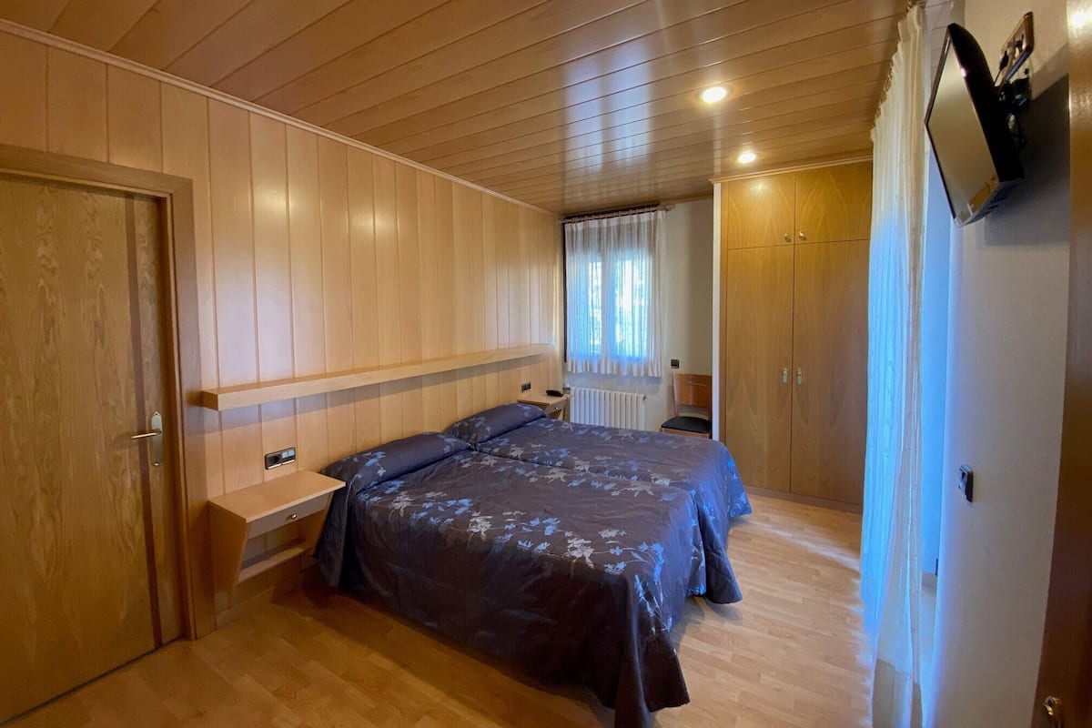 Quarto com paredes de madeira, cama com colcha floral, mesa de cabeceira, cadeira e guarda-roupa.