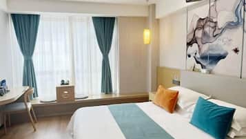 Quarto de hotel com cama, escrivaninha, cadeira, janela com cortinas e uma obra de arte na parede.