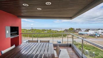 Varanda com mesa e cadeiras de madeira, vista para uma rua e vegetação.