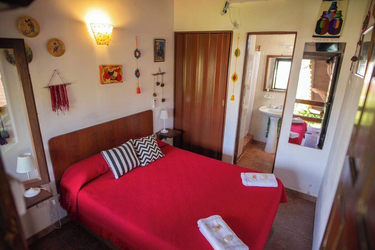 Quarto com colcha vermelha, cabeceira de madeira, abajur e luminária na parede.