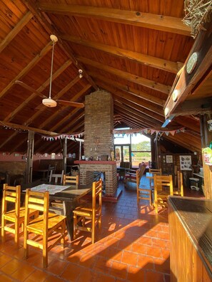 Interior rústico em madeira com lareira de tijolos, mesas e cadeiras de madeira, e piso ladrilhado.