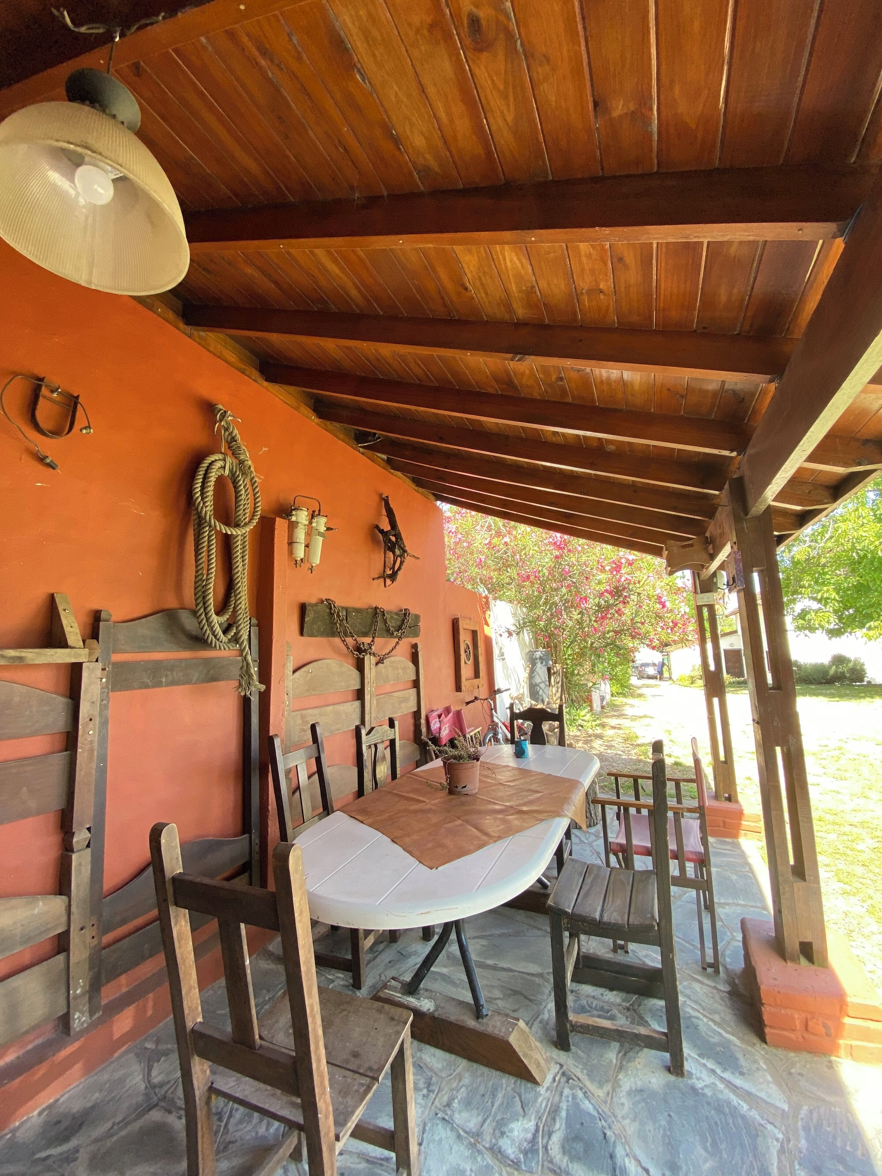 Uma pérgola de madeira ao ar livre com uma mesa e cadeiras, rodeada por piso de pedra e paredes laranja.