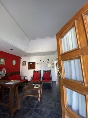 Quarto com sofá vermelho, duas cadeiras de madeira com almofadas vermelhas, uma mesa de madeira e uma porta de madeira com painéis de vidro.