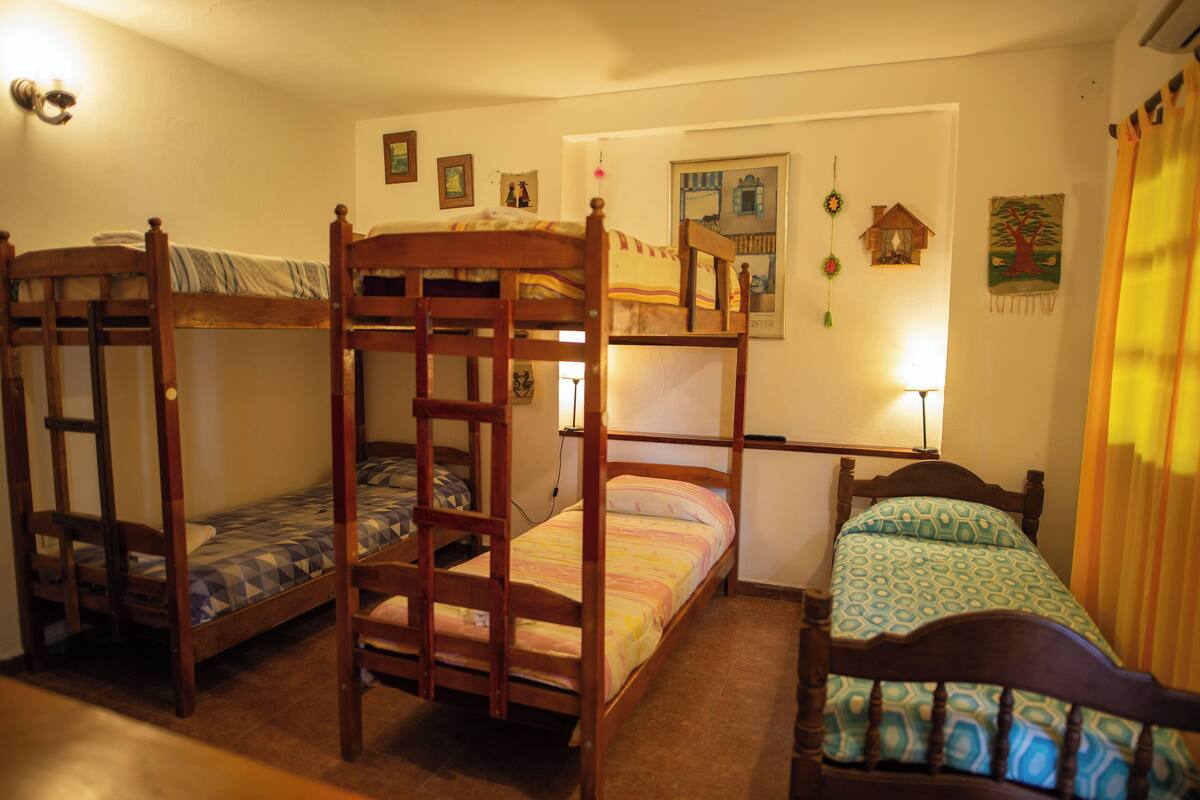Quarto com três beliches de madeira, uma mesinha e uma parede decorada com diversos objetos.