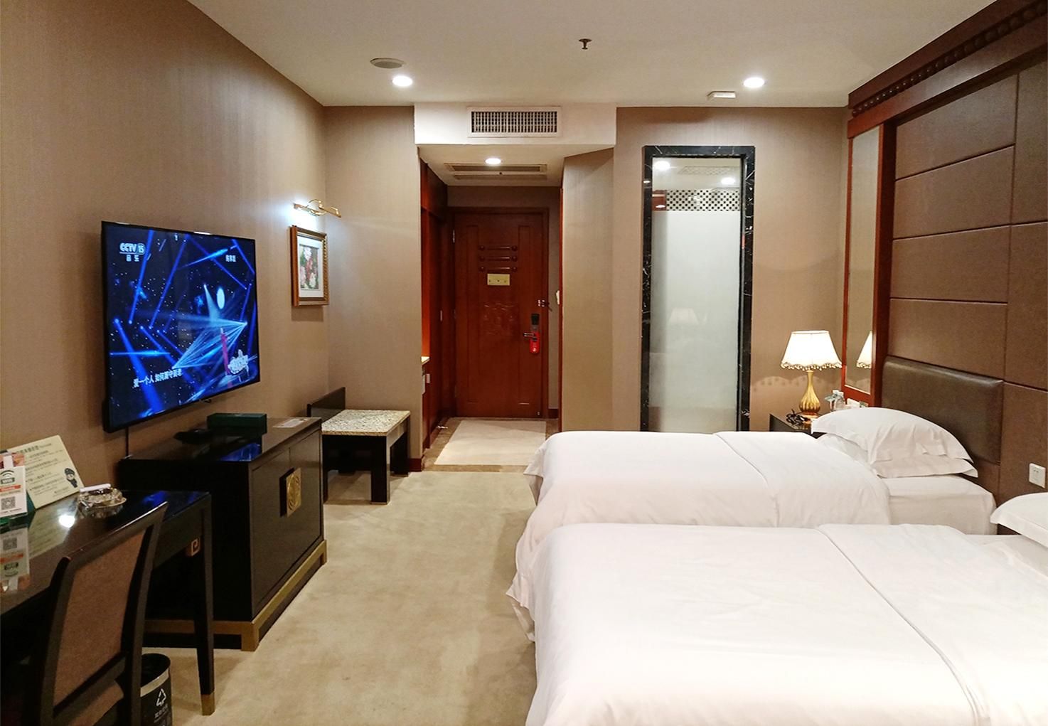 Quarto de hotel com televisão, secretária, duas camas, espelho e porta.