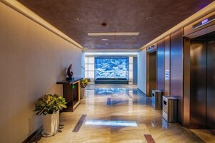 Um hall moderno de hotel com piso lustroso, balcão de recepção, plantas em vasos e elevadores.