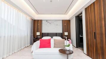 Quarto de hotel moderno com um sofá branco, uma mesa redonda pequena, uma cama com cabeceira e uma janela grande com cortinas leves.