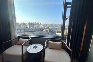 Quarto com duas cadeiras e uma pequena mesa, uma janela grande com vista para a cidade e uma cortina.