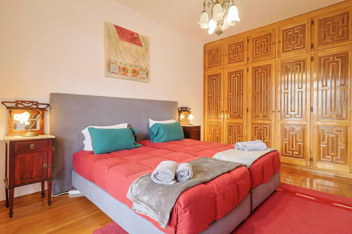 Uma cama bem arrumada com roupa de cama vermelha e duas almofadas, um guarda-roupa de madeira com detalhes elaborados e uma obra de arte na parede.