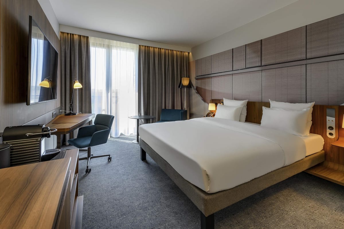 Quarto de hotel com uma cama grande, uma escrivaninha, uma cadeira, uma televisão e um abajur.