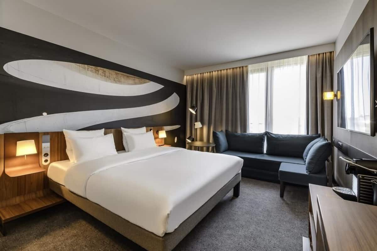 Quarto de hotel moderno com uma cama grande, um sofá, uma escrivaninha e uma televisão.