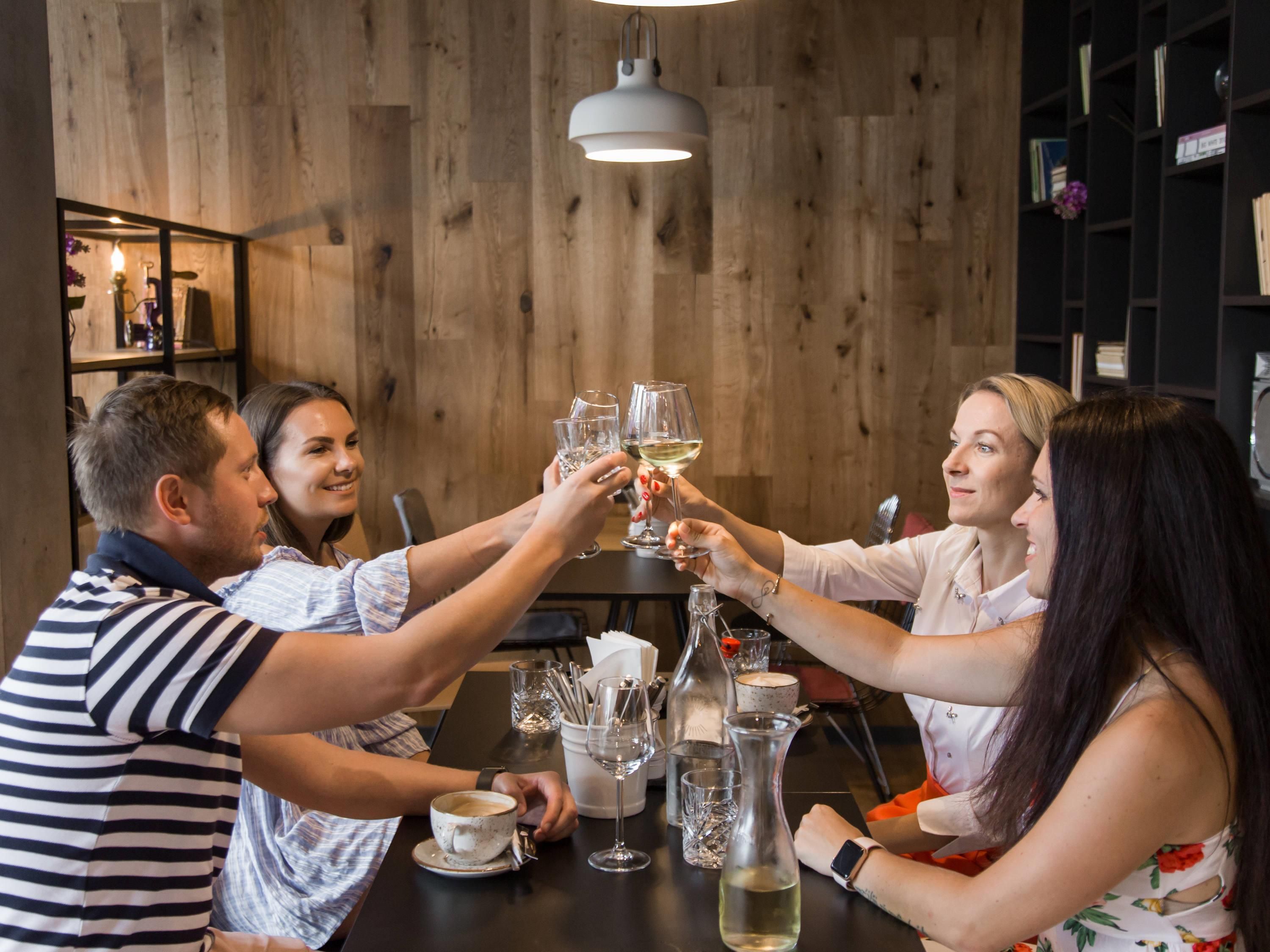 Um grupo de pessoas brindando com taças de vinho em uma mesa, em um ambiente com paredes de madeira e uma estante de livros.