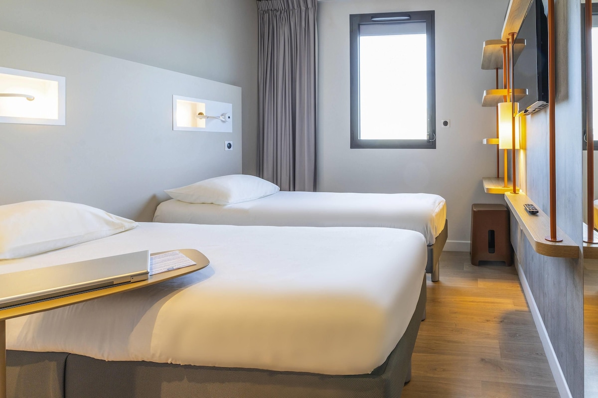 Quarto de hotel com duas camas, uma janela, uma mesa de cabeceira com abajur e um banquinho pequeno.