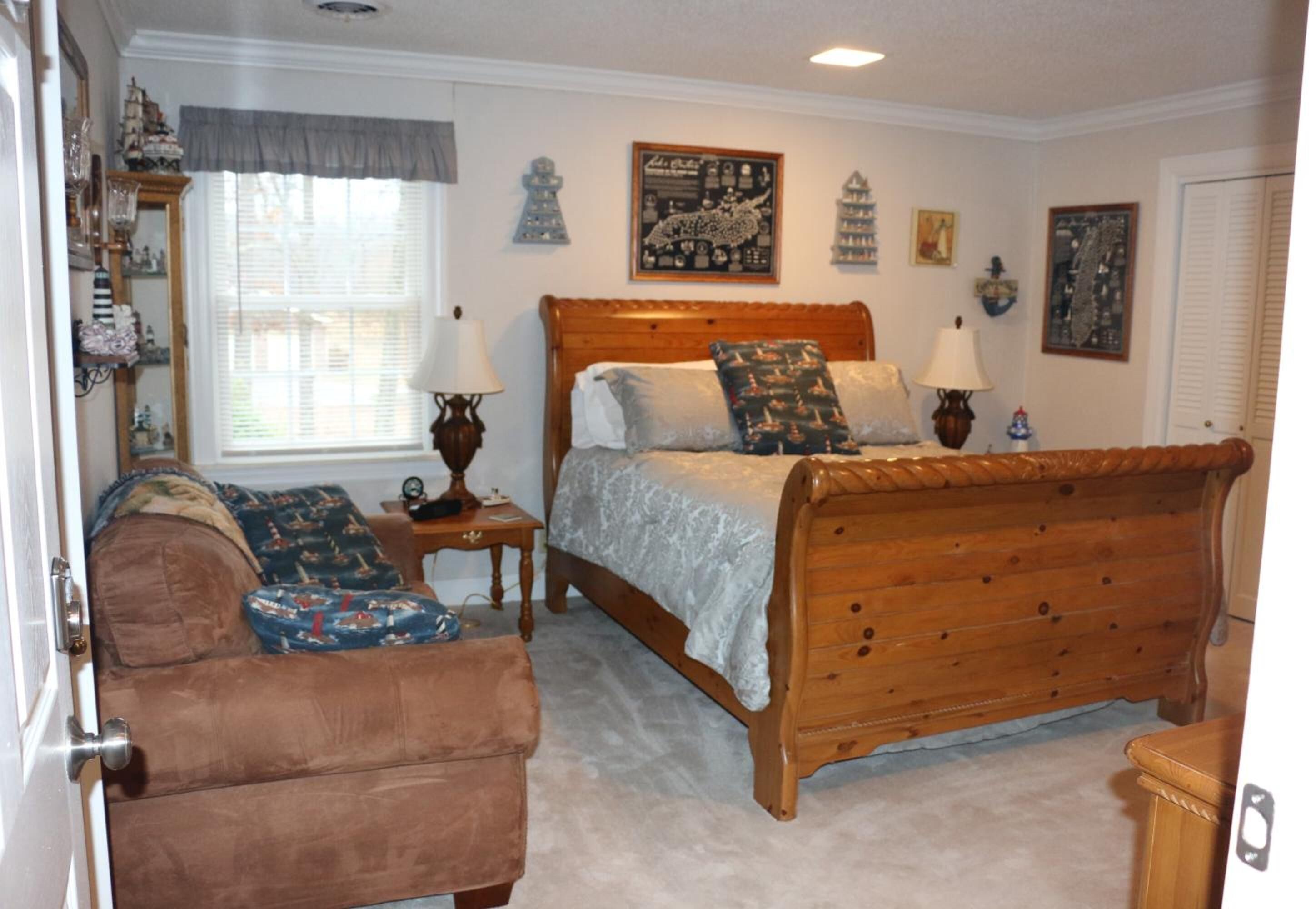 Quarto com cama de madeira, uma poltrona marrom, uma mesinha de cabeceira com abajur e uma janela com persianas.