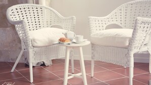 Duas cadeiras de vime brancas com almofadas, uma mesa redonda pequena e um prato com pastelaria e uma chávena.