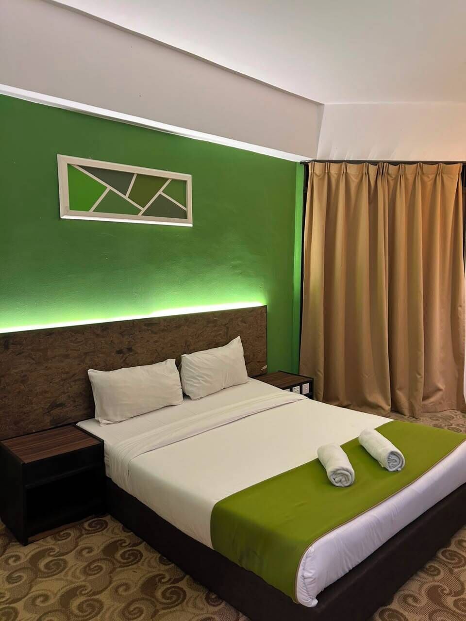 Quarto de hotel com cama, mesa de cabeceira, parede verde, decoração geométrica e cortina.