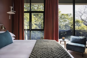 Quarto de hotel com uma cama grande, uma mesinha de cabeceira, uma cadeira e vista para árvores através de janelas amplas.