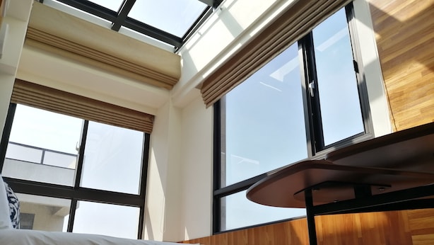 Quarto com janelas amplas, uma cama, uma mesa de madeira e um travesseiro verde com padrão.