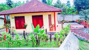 Uma casa térrea com telhado de telhas vermelhas, varanda com guarda-corpo vermelho e um pequeno jardim com diversas plantas.