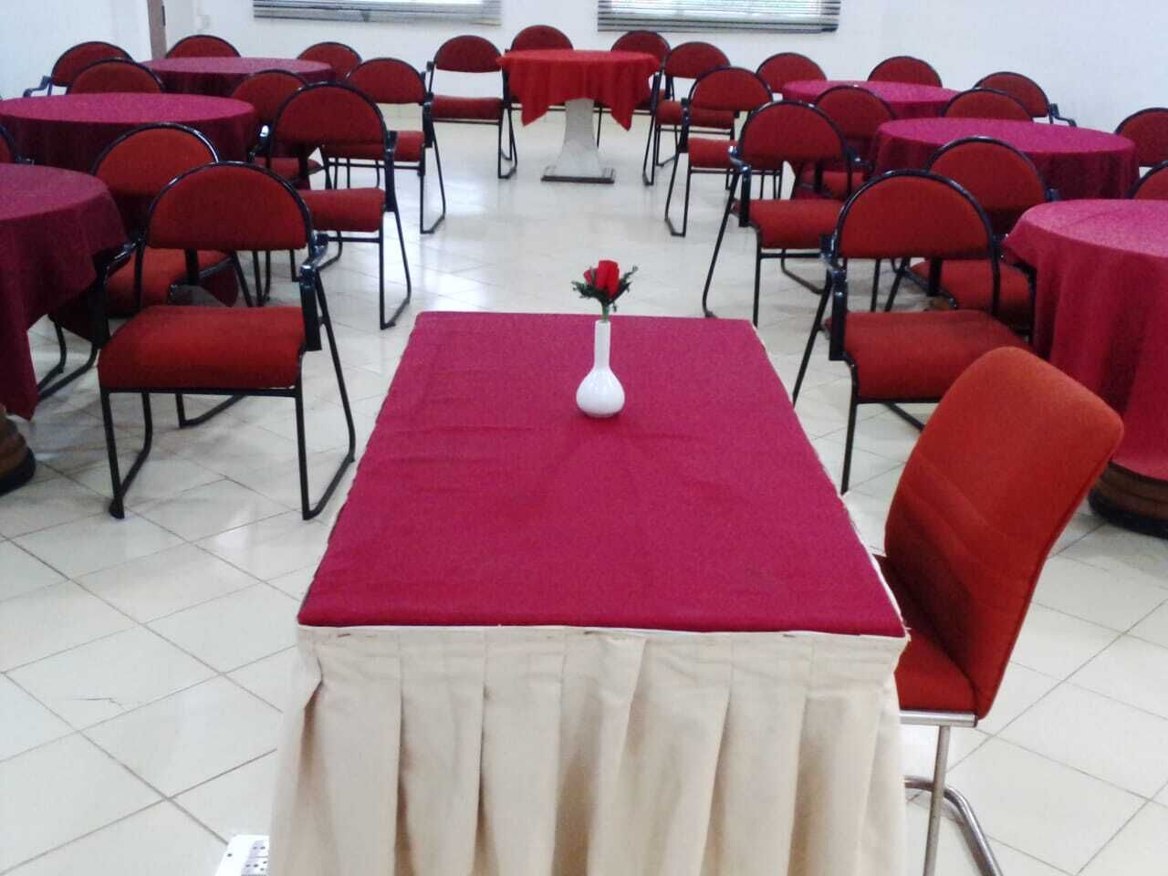 Uma sala de conferências com cadeiras e mesas vermelhas, um vaso branco com uma flor vermelha e um caminho de mesa bege.