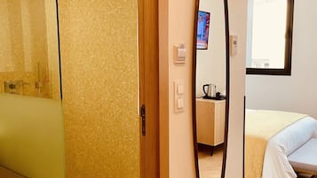 Quarto de hotel com box de vidro, porta de madeira, espelho grande, cama com colcha branca e amarela, mesa de cabeceira e janela com vista.