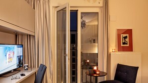 Quarto de hotel com escrivaninha, duas cadeiras, computador, televisão, porta de vidro deslizante, mesinha com velas e quadro na parede.