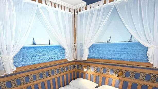 Quarto com cama, duas almofadas, colcha azul e vista para o mar com veleiros.