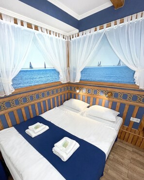Quarto com cama, duas almofadas, colcha azul e vista para o mar com veleiros.