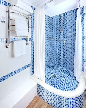 Casa de banho com chuveiro em azulejos azuis, banheira branca e suporte de toalhas com toalhas dobradas.