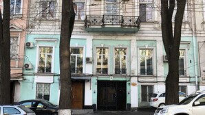 Um edifício de vários andares com fachada verde, entrada central com portão preto e carros estacionados na rua.