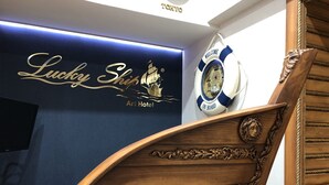 Uma escadaria de madeira com esculturas detalhadas, uma placa com a inscrição "Lucky Ship Art Hotel" e um círculo salva-vidas com a frase "BEM-VINDO A BORDO" na parede.