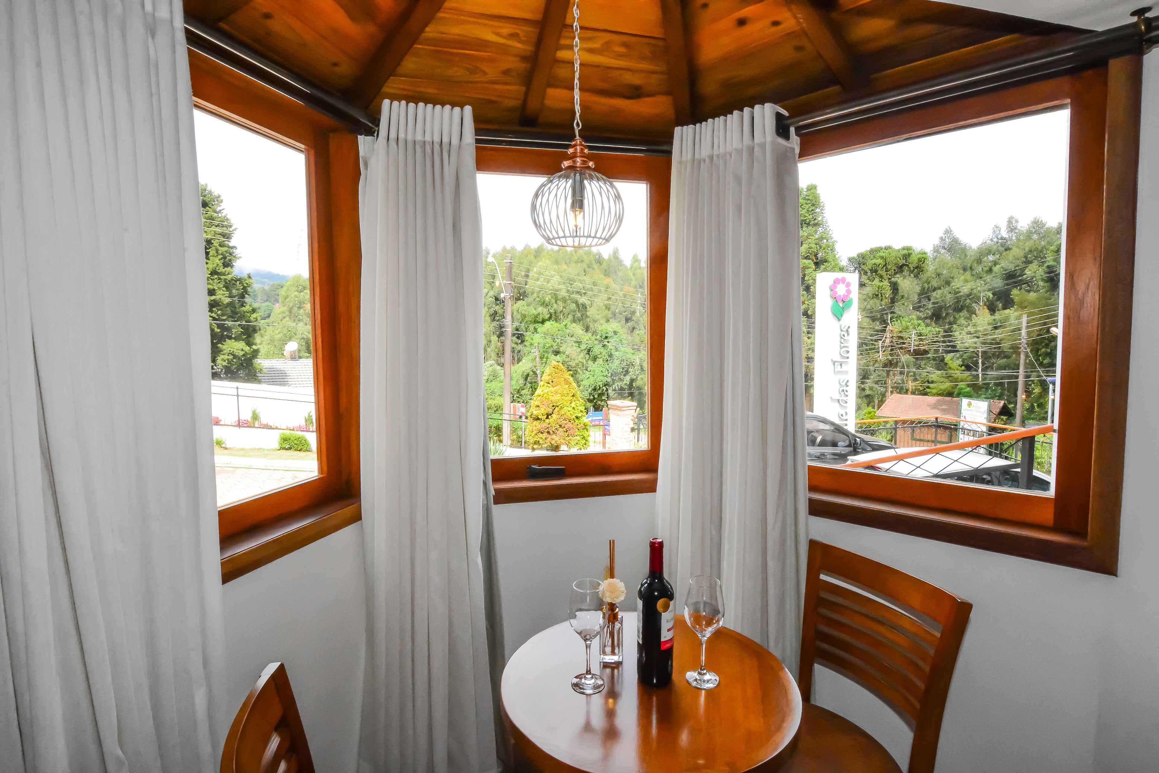 Mesa de jantar de madeira com uma garrafa de vinho e duas taças, em frente a uma janela com cortinas brancas.