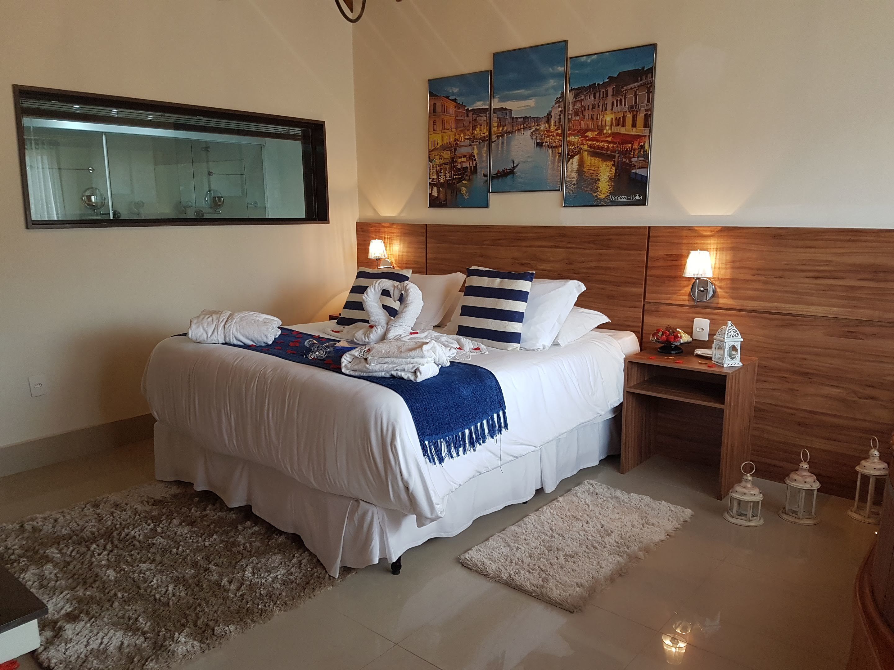 Quarto de hotel com uma cama grande, cabeceira de madeira, mesa de cabeceira com abajur e espelho fixado na parede.