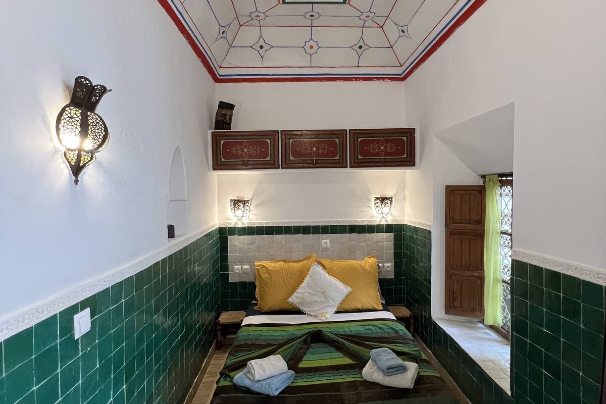 Quarto pequeno com uma cama, paredes com azulejos verdes, teto estampado e uma janela com cortina.