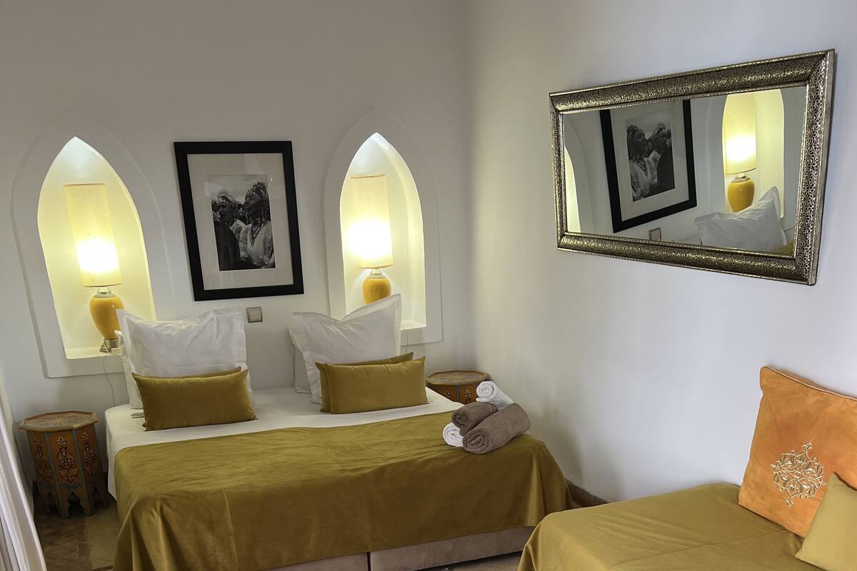 Quarto de hotel com duas camas, um tapete vermelho e branco estampado e um banquinho amarelo.